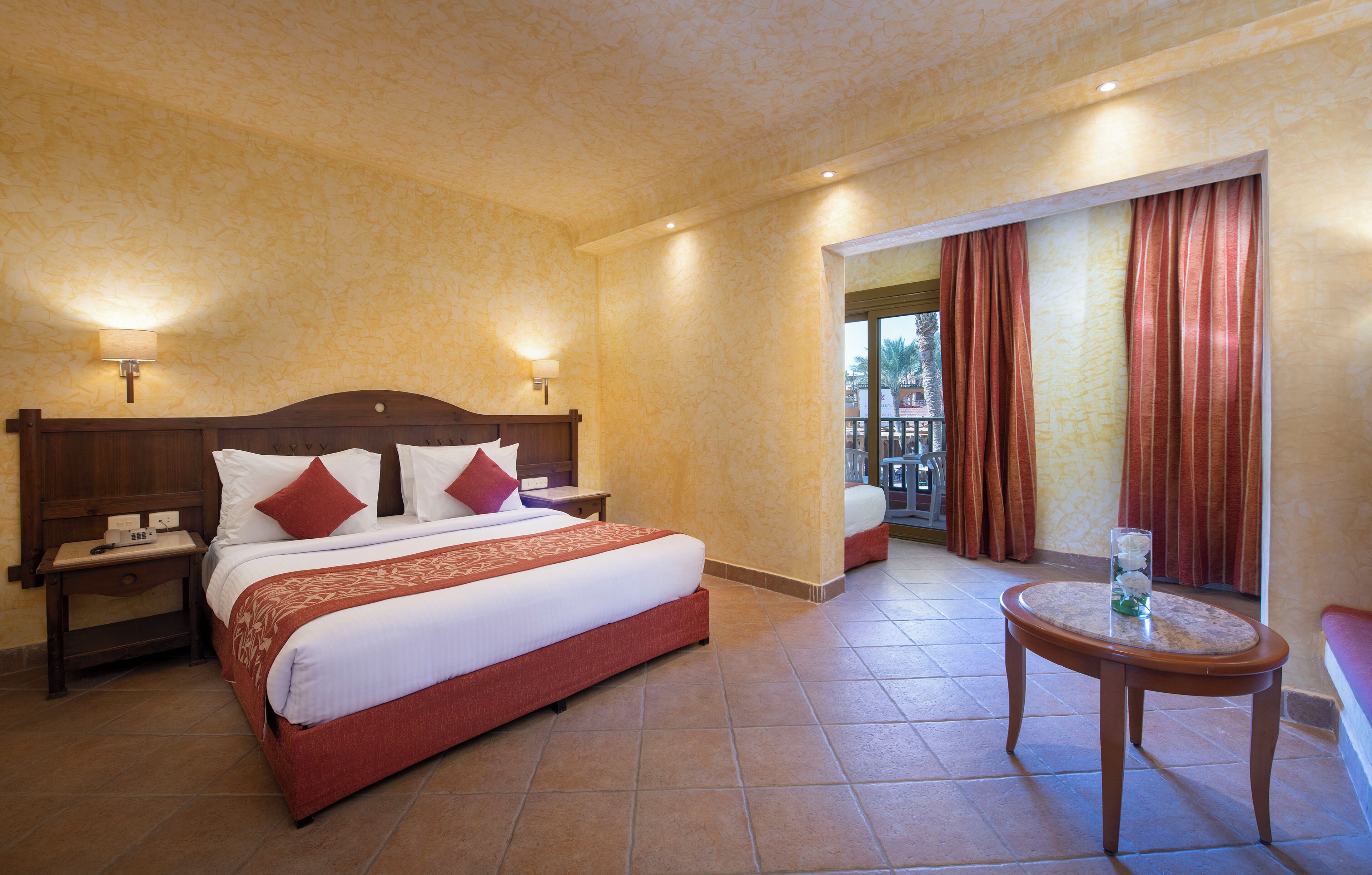 Quarto de hotel com uma cama grande, duas mesas de cabeceira, uma mesa redonda e vista para o exterior.