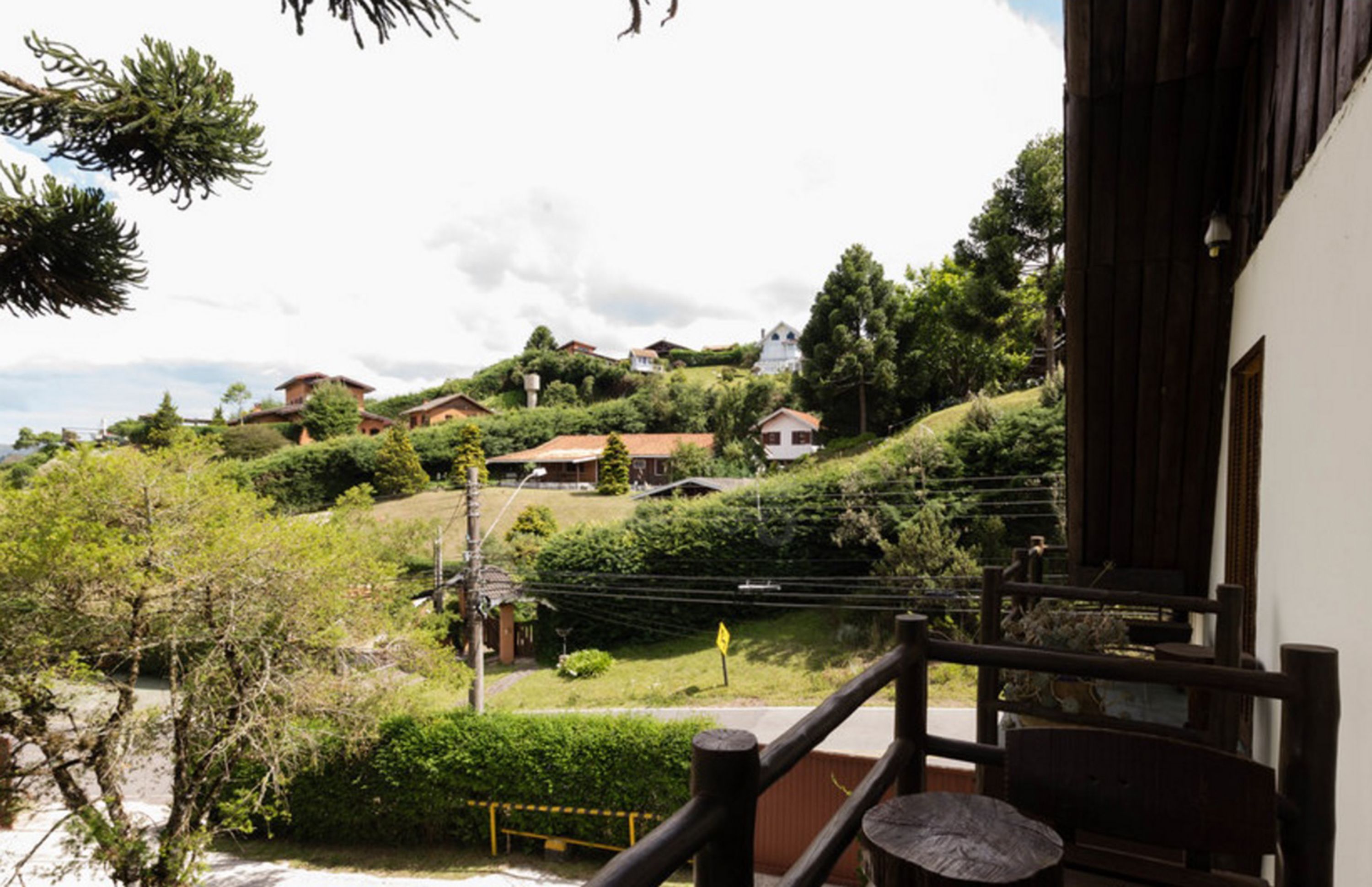 Vista de uma varanda com vista para uma encosta onde se veem algumas casas, um cercado de madeira e um céu claro.