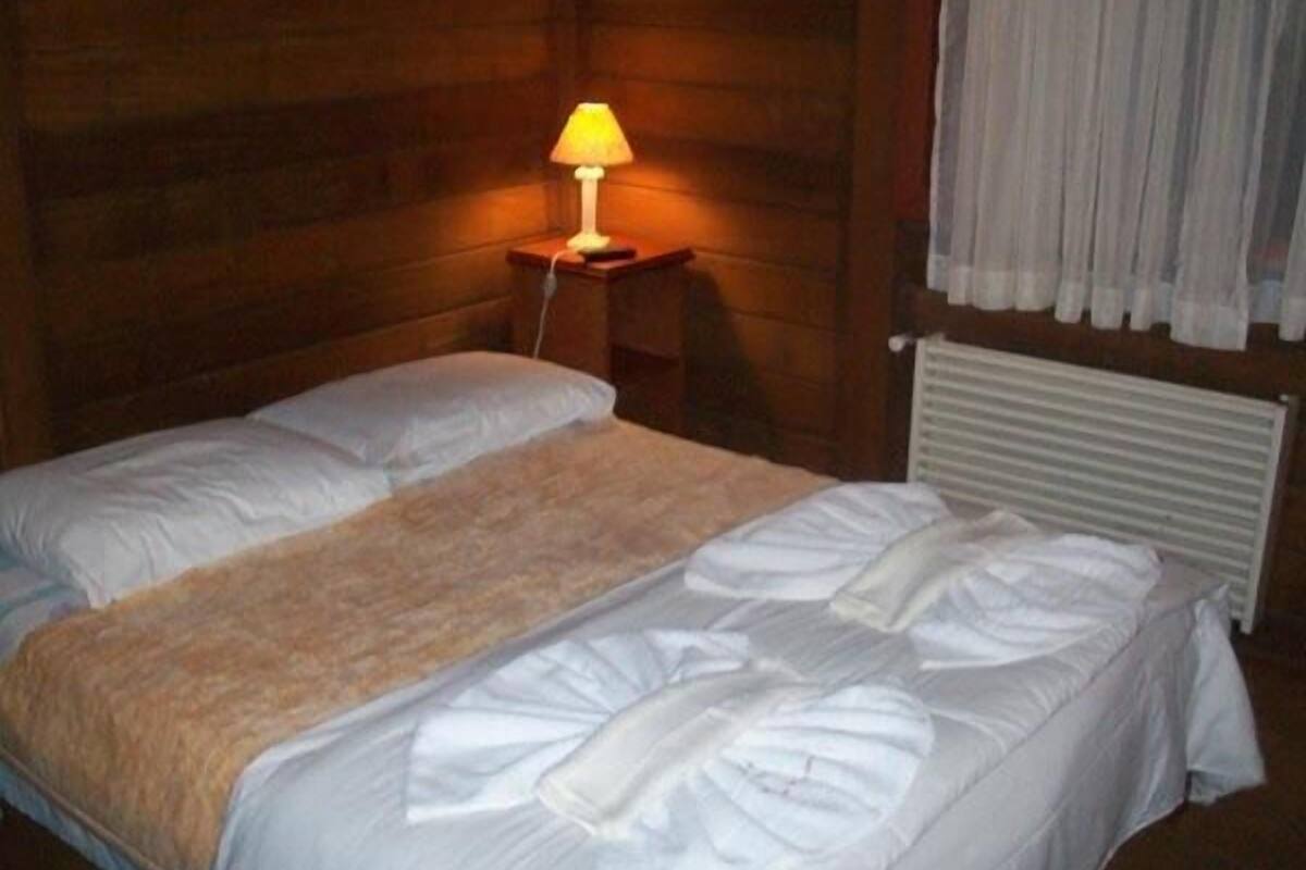 Cama de solteiro com roupa de cama branca e um cobertor bege num quarto com paredes de madeira e radiador branco.