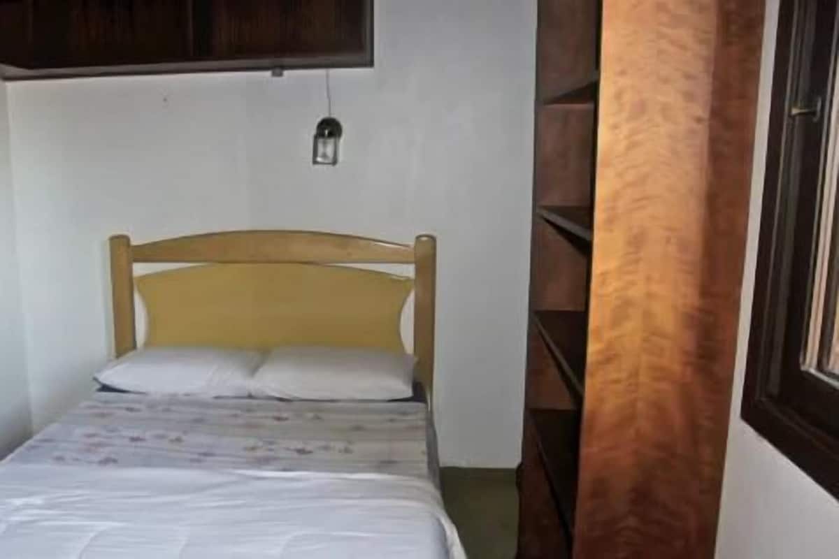 Quarto pequeno com uma cama de solteiro, cabeceira de madeira, colcha branca e luminária pequena fixada na parede.