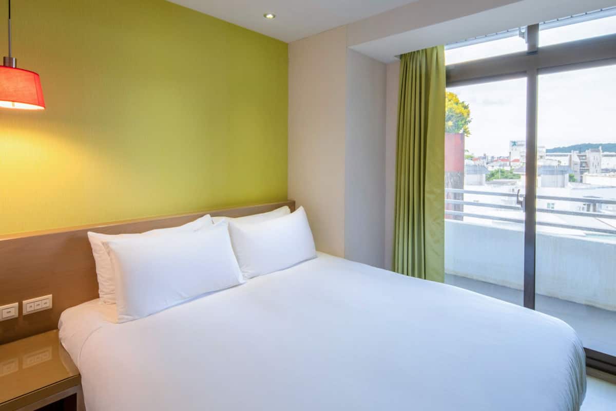 Quarto de hotel com uma cama, mesa de cabeceira, abajur vermelho, cortina verde e vista para a cidade pela janela.