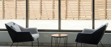 Duas cadeiras pretas com almofadas cinzentas, uma mesa redonda pequena e vista para uma cerca de madeira através de janelas amplas.