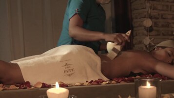 Uma pessoa deitada em uma cama com pétalas de rosas, uma vela acesa e uma toalha com texto.
