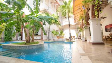 Piscina interior com borda azulejada, rodeada por vegetação exuberante e um edifício de vários níveis com varandas.