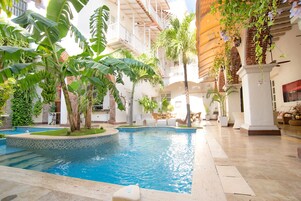 Piscina interior com borda azulejada, rodeada por vegetação exuberante e um edifício de vários níveis com varandas.