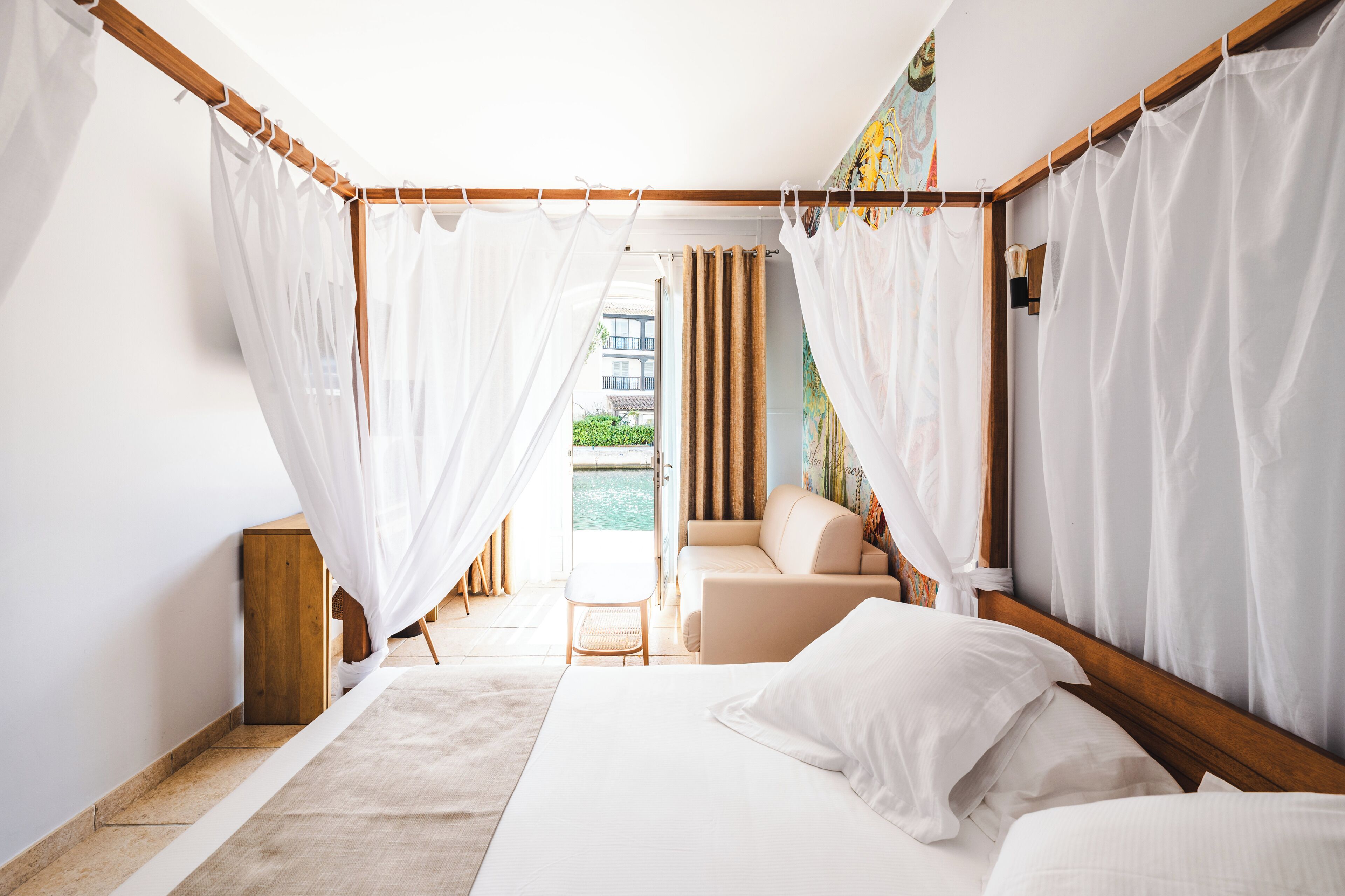 Quarto com cama, cabeceira de madeira, mesa de cabeceira de madeira, sofá bege e vista para o exterior.