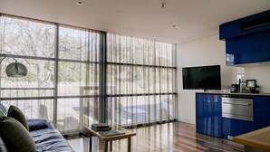 Uma sala de estar moderna com sofá, mesa de centro e televisão. Há uma cozinha com armários azuis e fogão.