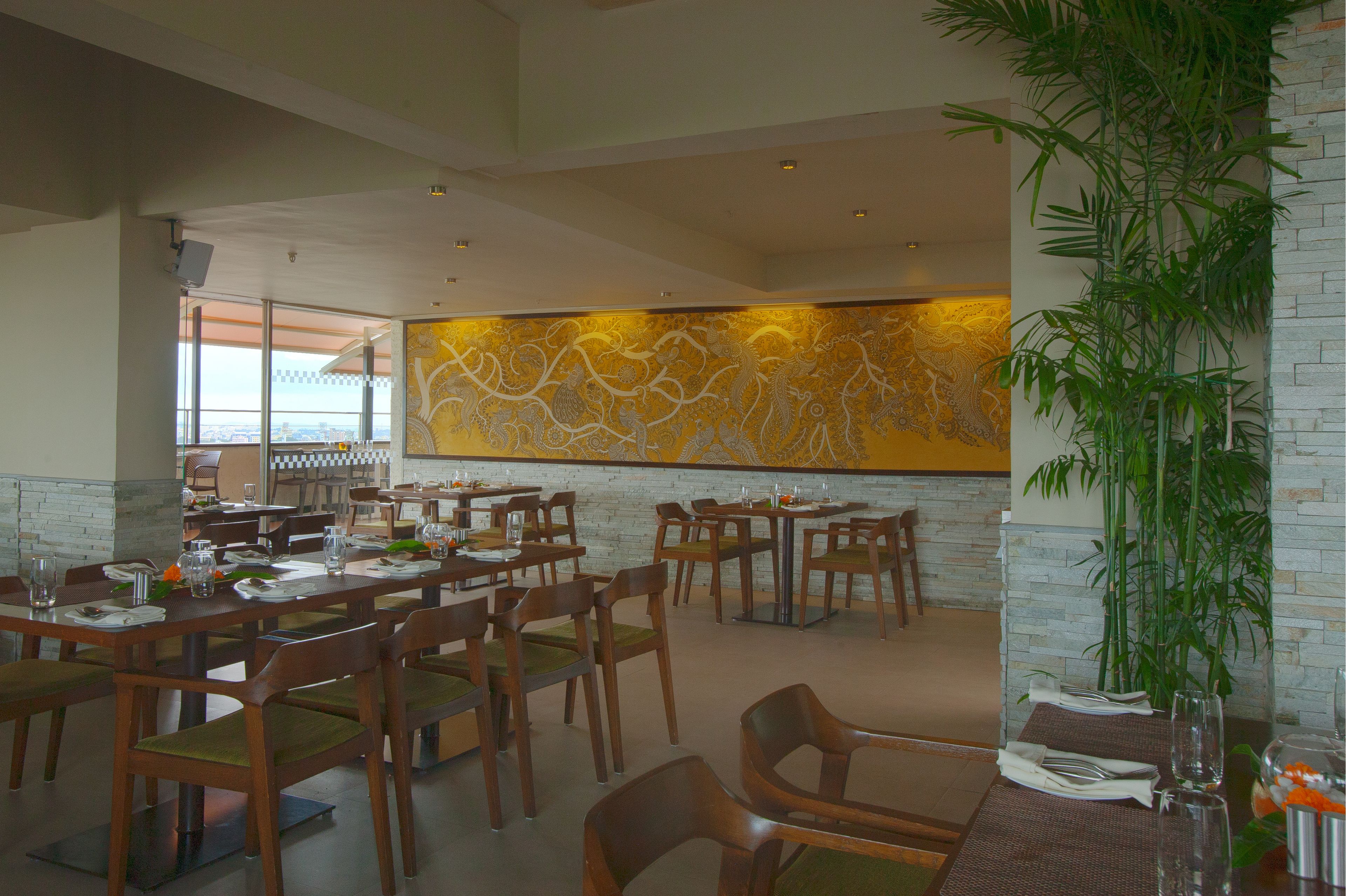 Um restaurante com mesas e cadeiras de madeira, um painel decorativo grande e uma planta de bambu.