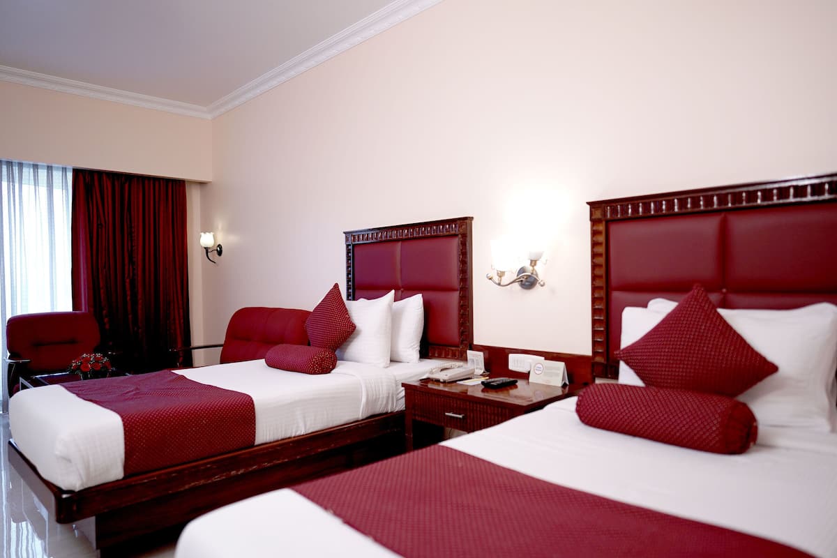 Quarto de hotel com duas camas, cada uma com roupa de cama e almofadas vermelhas, cabeceira de madeira, mesa de cabeceira com abajur e uma cadeira vermelha.