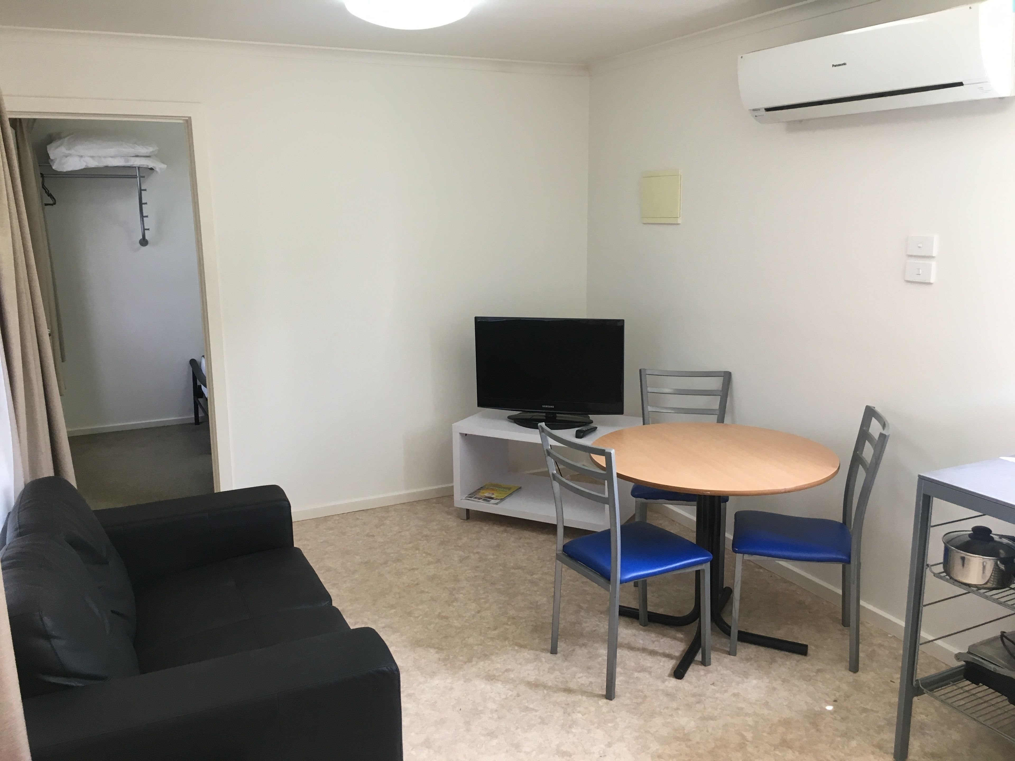 Um espaço compacto com um sofá preto, uma mesinha redonda com duas cadeiras azuis, uma TV sobre um suporte branco e um ar condicionado branco na parede.