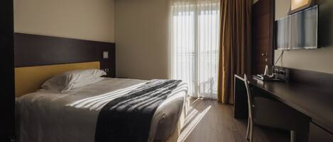 Quarto de hotel com cama, escrivaninha, cadeira, televisão e janela com cortinas.