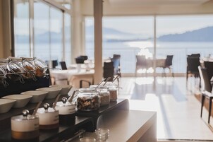 Uma mesa de pequeno-almoço com potes de granola e acompanhamentos, com vista para uma paisagem deslumbrante de um corpo de água e montanhas.