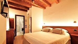 Quarto com teto de madeira, cama com roupa de cama bege, mesa de cabeceira e escrivaninha de madeira com cadeira.