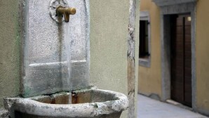 Uma fonte de água ao ar livre desgastada pelo tempo, com um bico e uma bacia de metal, posicionada diante de uma parede texturizada com grafite.