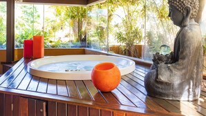 Uma banheira de hidromassagem com uma estátua de Buda, cercada por plantas e uma janela com vista para o verde.