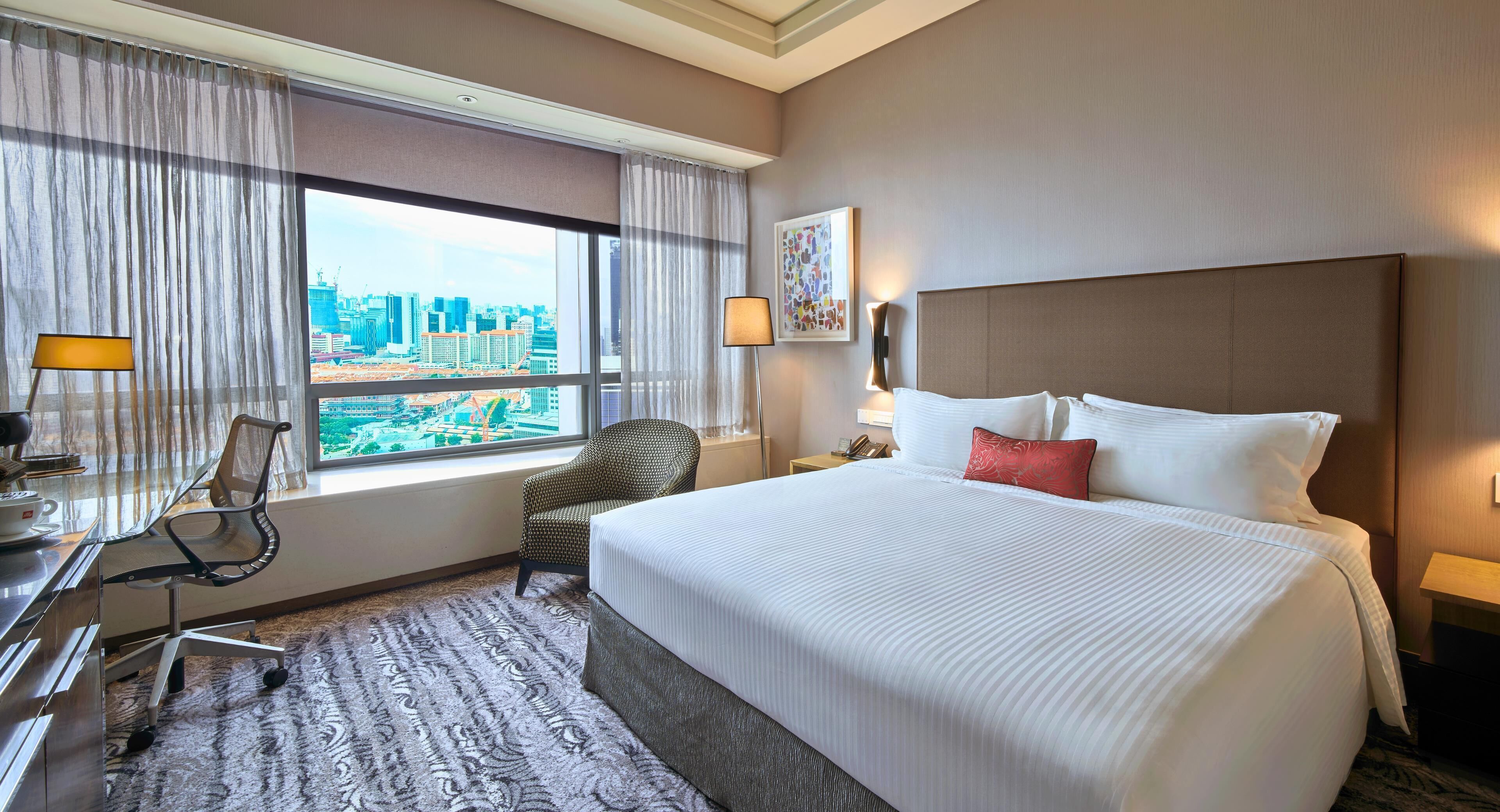 Quarto de hotel com cama grande, escrivaninha com cadeira, vista da cidade pela janela e abajur na mesa de cabeceira.