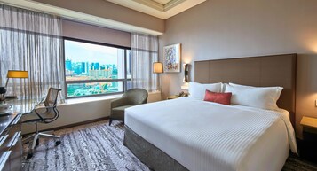 Quarto de hotel com cama grande, escrivaninha com cadeira, vista da cidade pela janela e abajur na mesa de cabeceira.