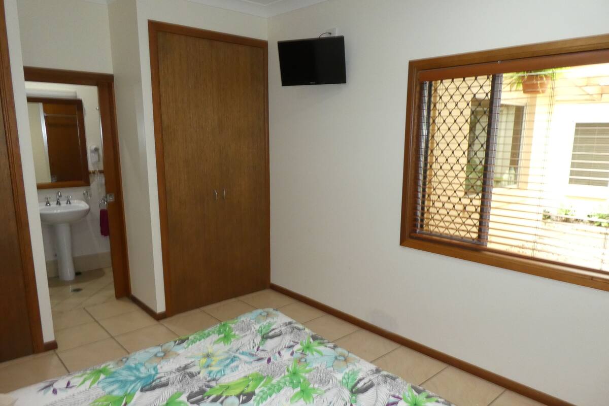 Quarto com cama, guarda-roupa, televisão fixada na parede e janela com vista para o exterior.