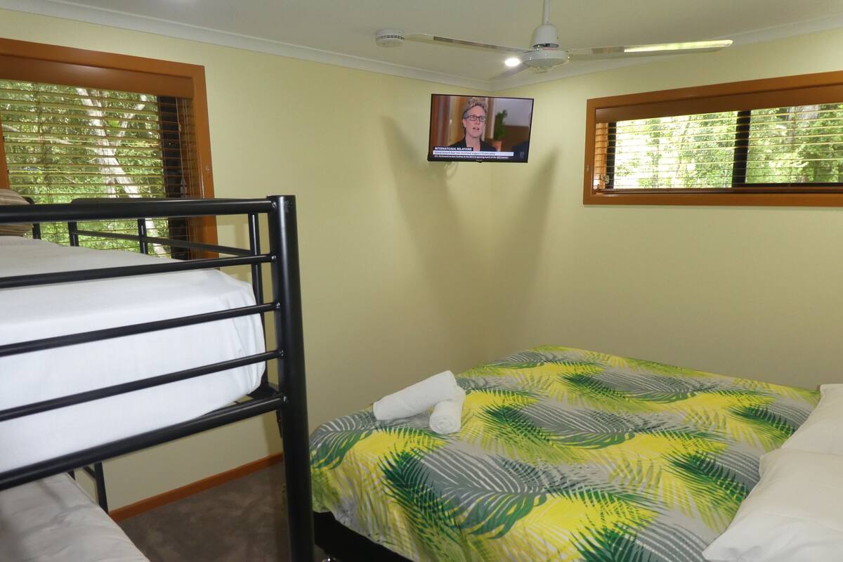Quarto com beliches, televisão fixada na parede e janela com persianas.