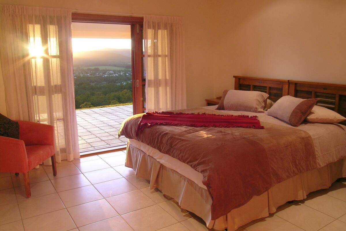 Quarto com uma cama grande, uma cadeira e vista para a paisagem através de uma porta aberta.