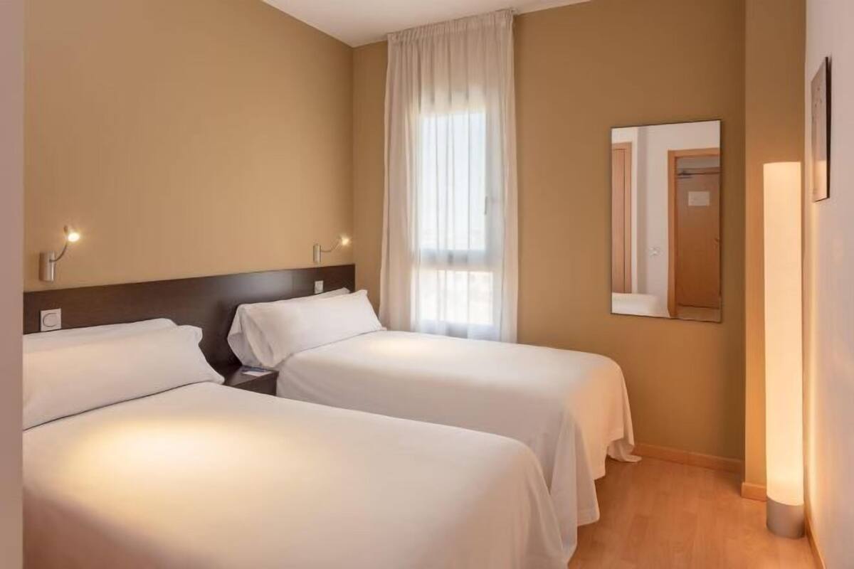Quarto de hotel com duas camas, uma mesa de cabeceira, janela com cortinas, espelho e luminária na parede.
