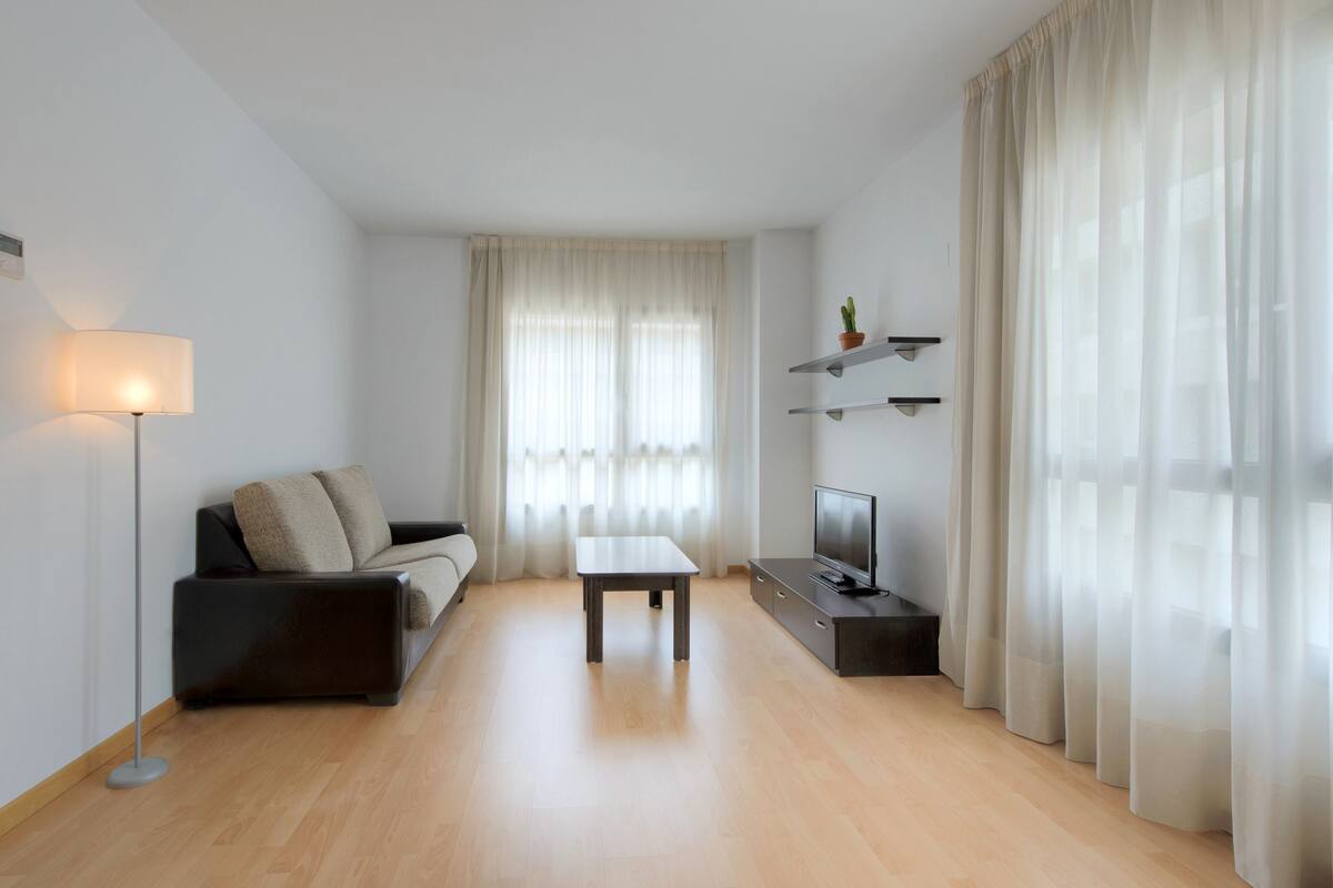 Uma sala de estar moderna com um sofá, uma mesa de centro, uma TV em um móvel e cortinas leves.