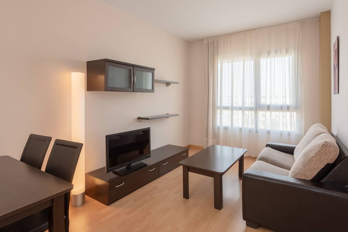 Uma sala de estar moderna com televisão de tela plana, móveis de madeira escura, um sofá e uma janela grande com cortinas leves.