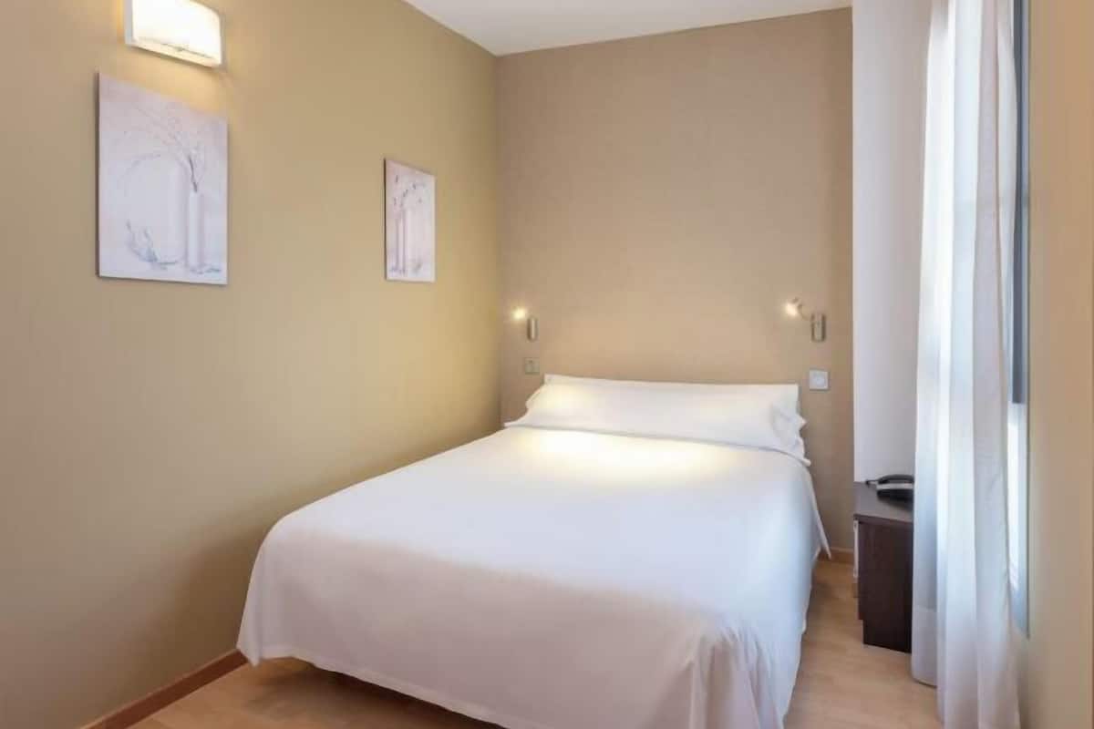 Uma cama de solteiro num quarto com paredes bege, duas luminárias fixadas na parede e uma janela com cortinas.