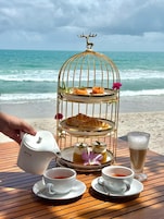 Uma mesa à beira-mar com um suporte escalonado para servir, duas chávenas de chá, um copo de café gelado e uma chaleira.