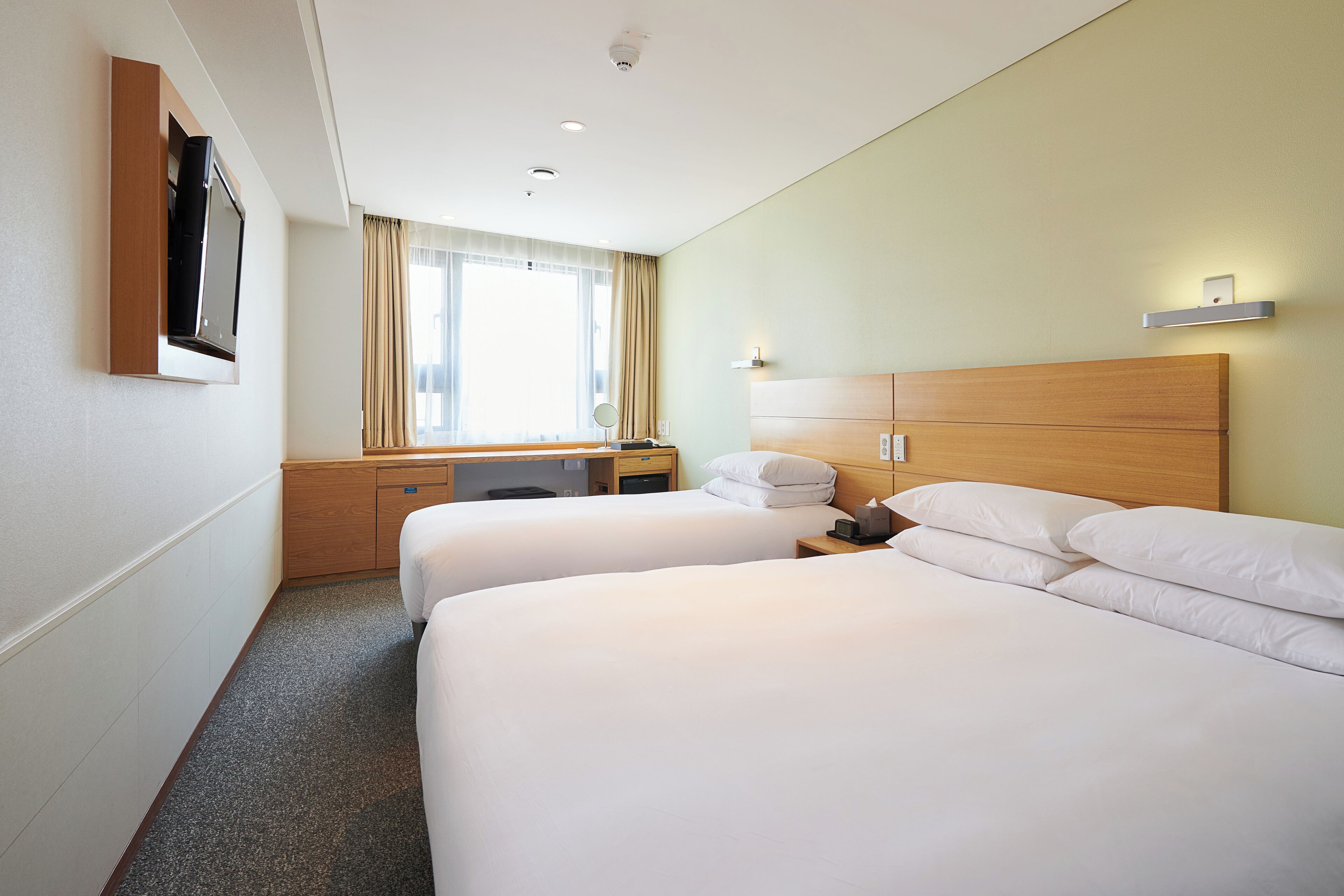 Quarto de hotel com duas camas, uma escrivaninha, televisão na parede e uma janela com cortinas.