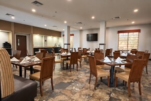 Uma sala de jantar espaçosa com várias mesas e cadeiras, área de cozinha e uma TV de tela plana na parede.