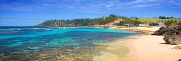 Uma praia costeira com águas turquesa cristalinas, areal arenoso e uma colina arborizada ao fundo.