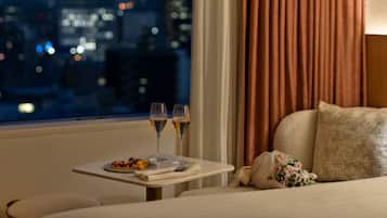 Quarto de hotel com vista para a cidade, uma mesa de jantar com um prato de comida e duas taças de vinho, e uma cama com travesseiro e um buquê de flores.
