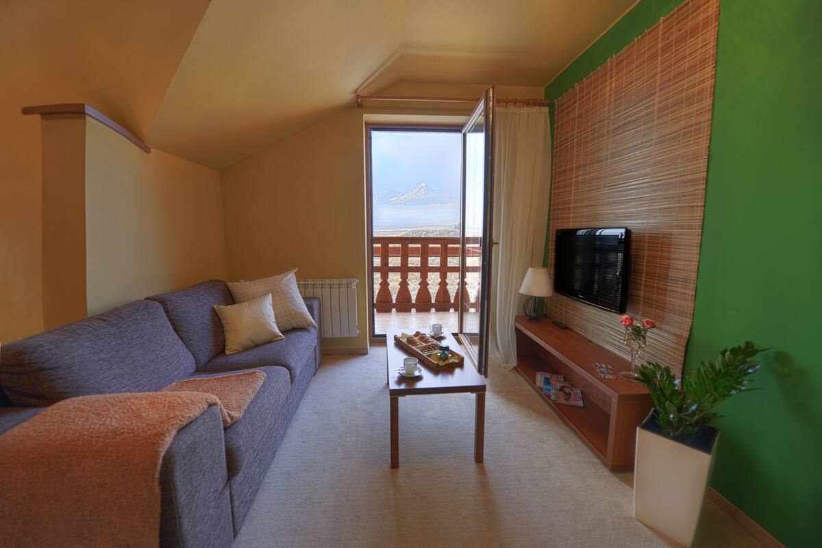 Sala de estar com sofá, mesa de centro, televisão e vista para uma varanda com montanhas ao fundo.