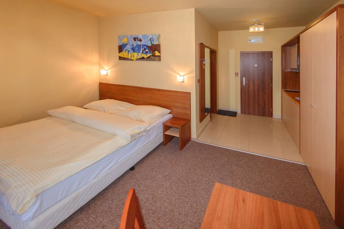 Quarto de hotel com uma cama de solteiro, cabeceira de madeira, mesa de cabeceira, luminária na parede, um quadro e uma porta que dá para um corredor.
