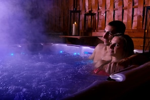 Um casal em uma banheira de hidromassagem, cercado por paredes de madeira e uma vela.