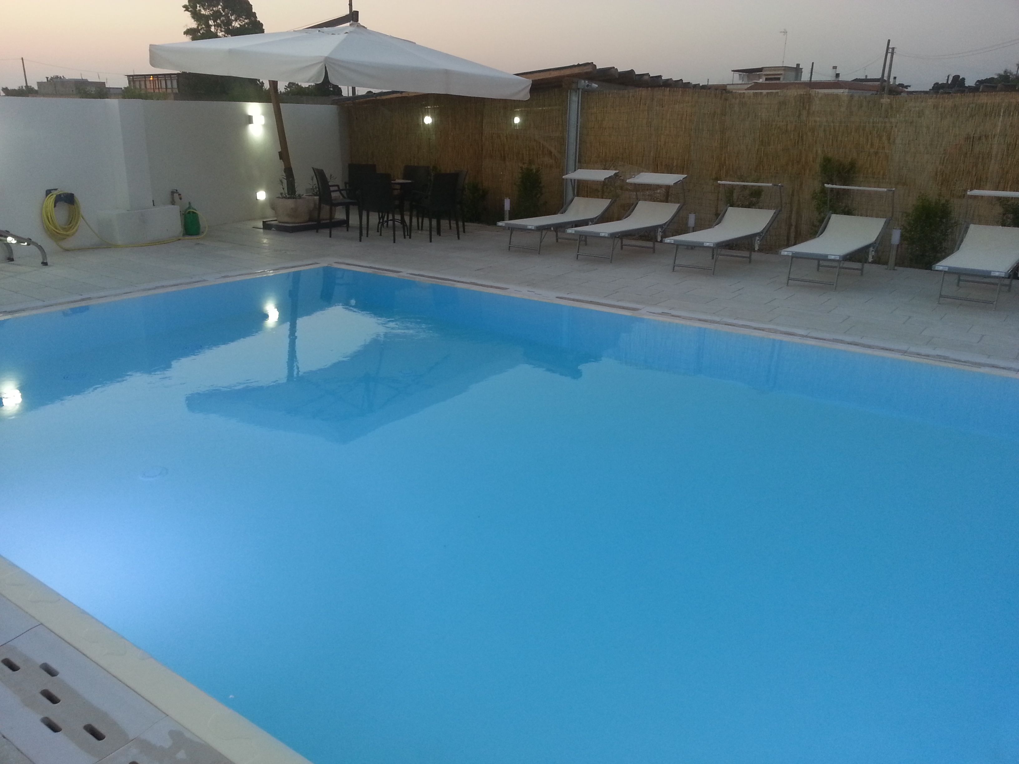Piscina com cadeiras de praia e guarda-sóis, cercada por um muro de madeira e uma parede branca.