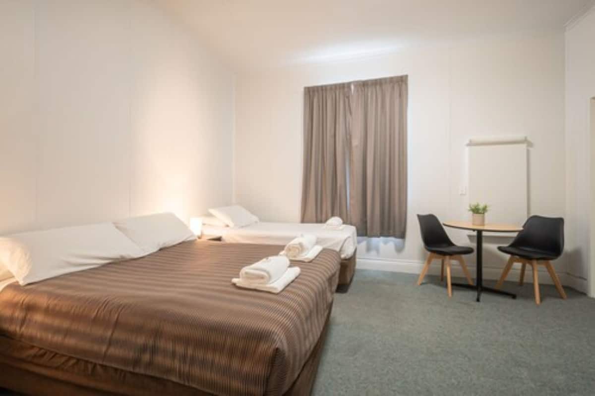 Quarto de hotel com duas camas, uma mesinha com cadeiras, um quadro branco e uma janela com cortinas.