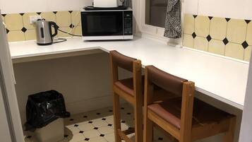 Cozinha compartilhada com micro-ondas, chaleira e lixeira. Há duas cadeiras de madeira e uma janela com cortinas xadrez.