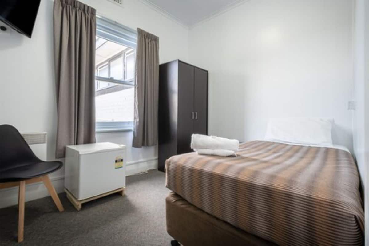Quarto de hotel com cama, guarda-roupa, escrivaninha, cadeira e janela com cortinas.