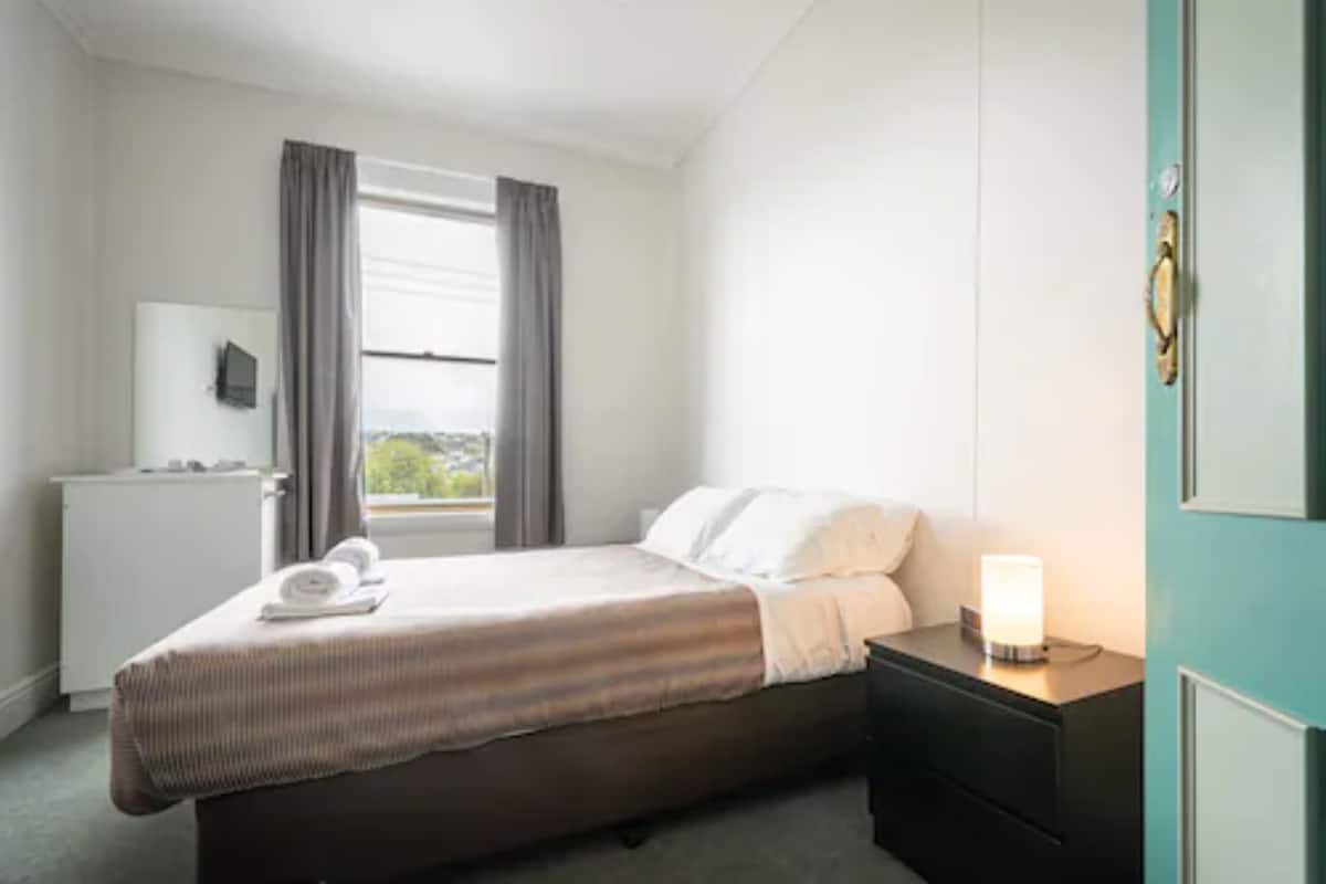 Quarto de hotel com cama, mesa de cabeceira com abajur, televisão em móvel e janela com cortinas.