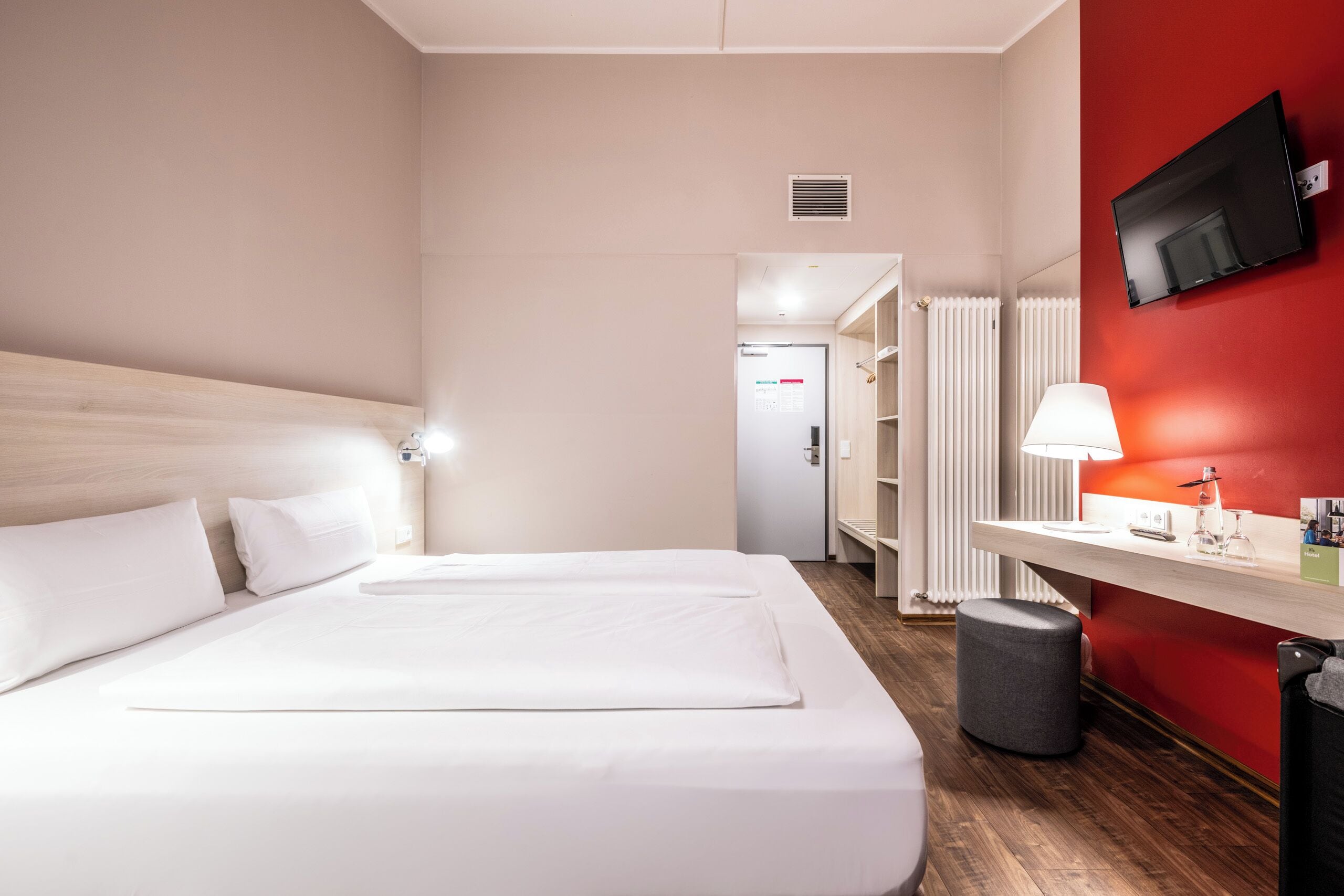 Quarto de hotel com cama, televisão na parede, mesa de cabeceira com abajur e um banquinho.