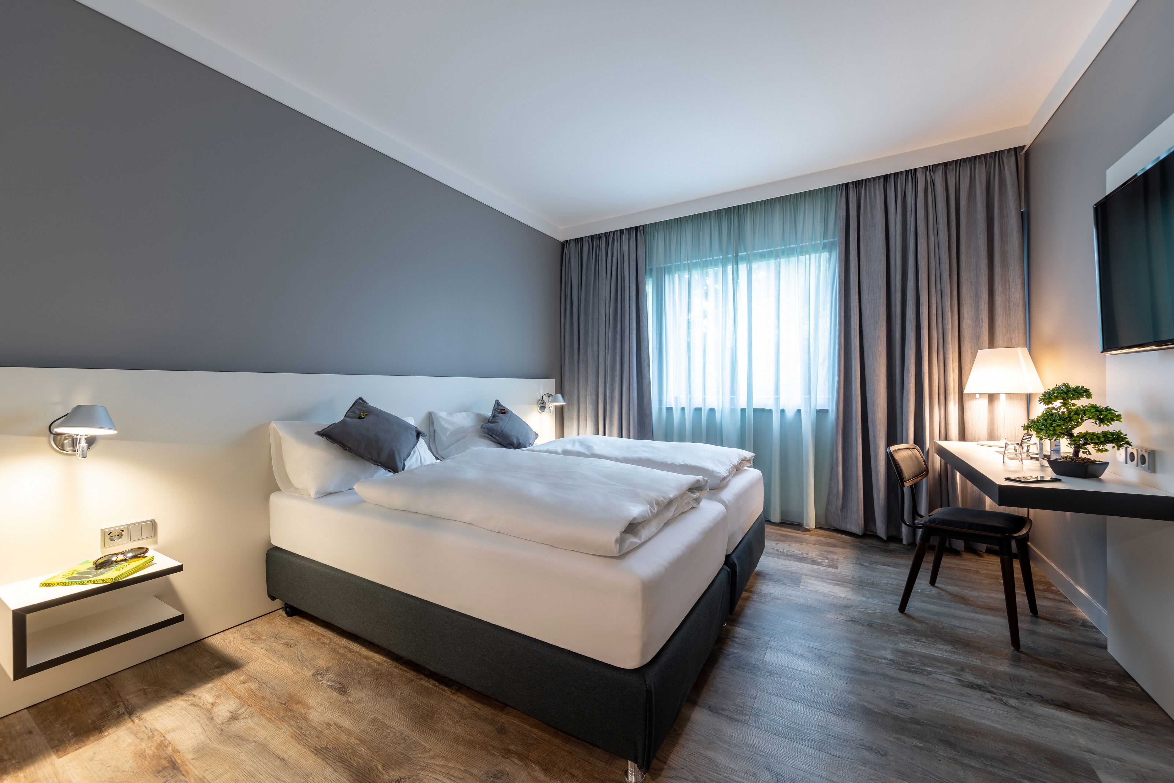 Quarto de hotel moderno com uma cama grande, mesas de cabeceira, escrivaninha e televisão.