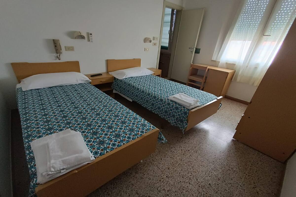 Quarto de hotel com duas camas, cada uma com uma colcha estampada e almofadas brancas. Há mesas de cabeceira com abajures e um pequeno armário.