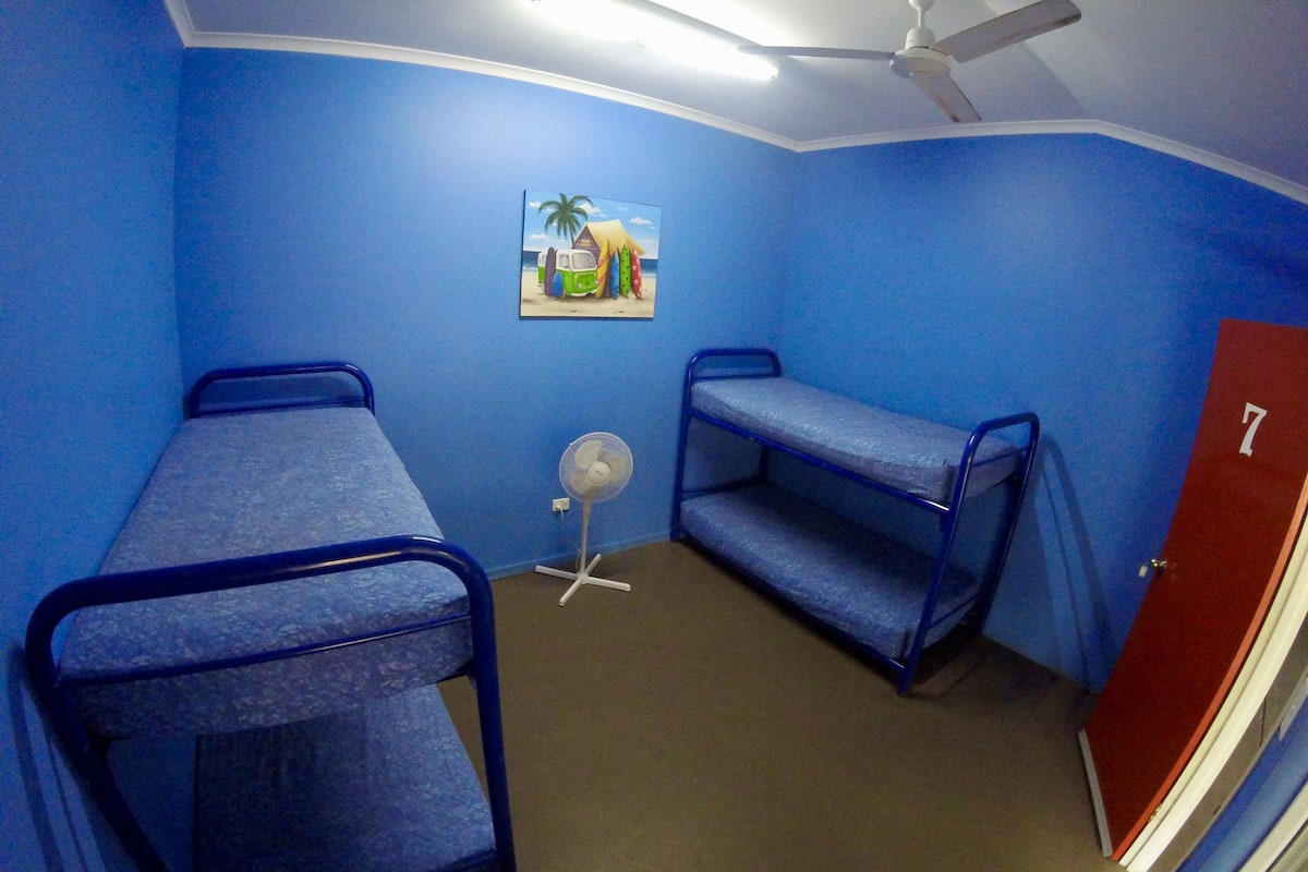 Quarto com duas camas beliche, um ventilador de teto, um ventilador fixado na parede e uma porta com o número 7.