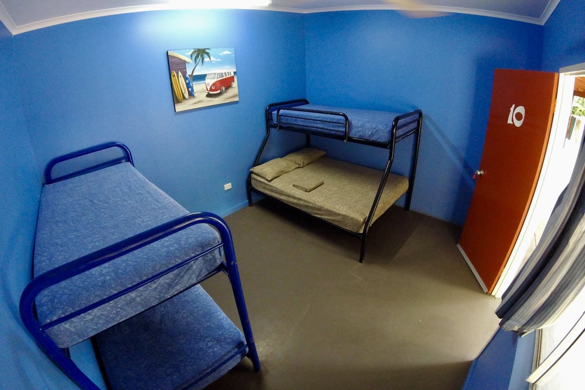 Um dormitório com duas beliches, uma cama de solteiro, um ventilador de teto, uma porta com o número 10 e um quadro na parede.