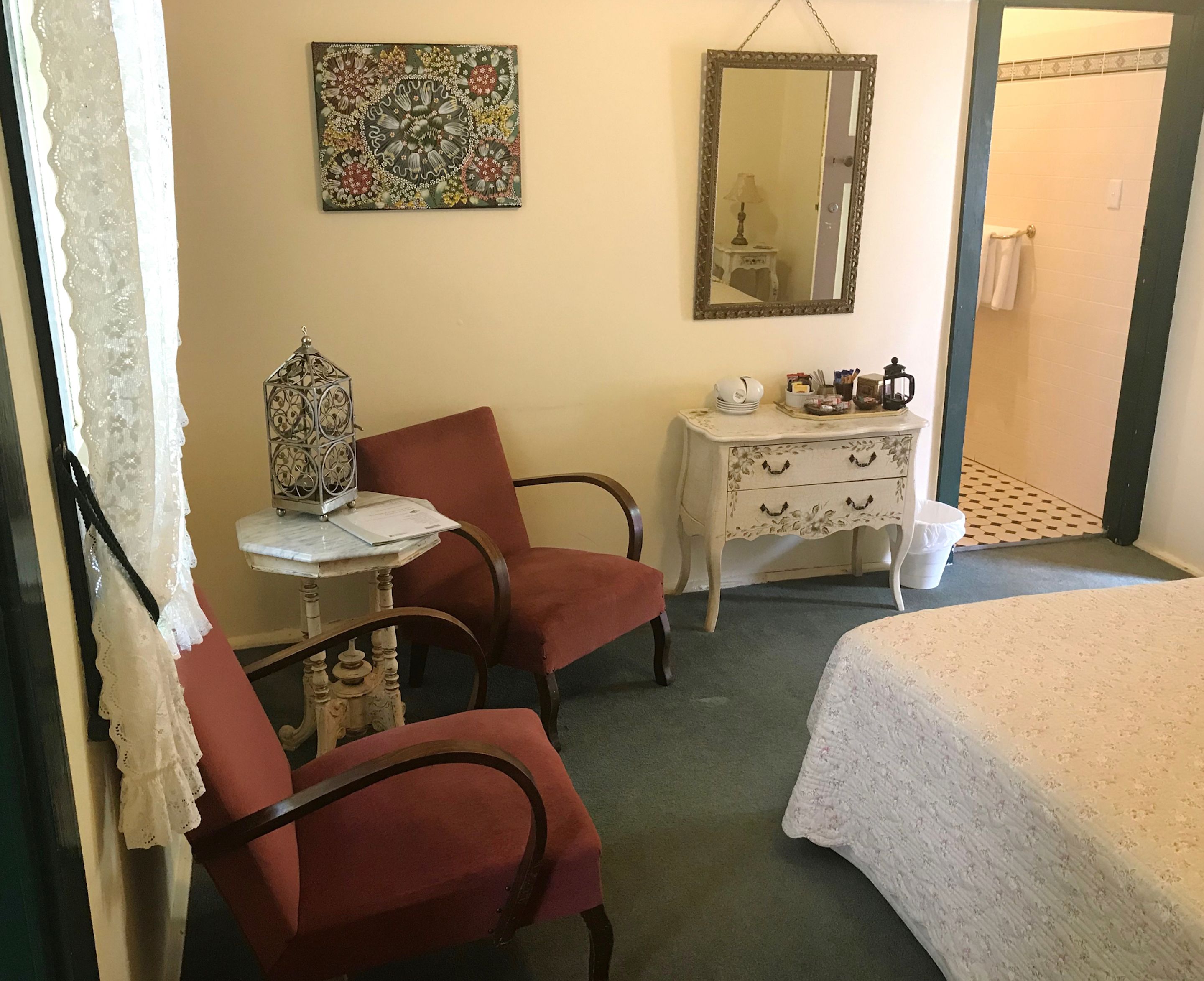 Quarto com cama, uma cadeira vermelha, mesa pequena com abajur, cômoda e espelho fixado na parede.