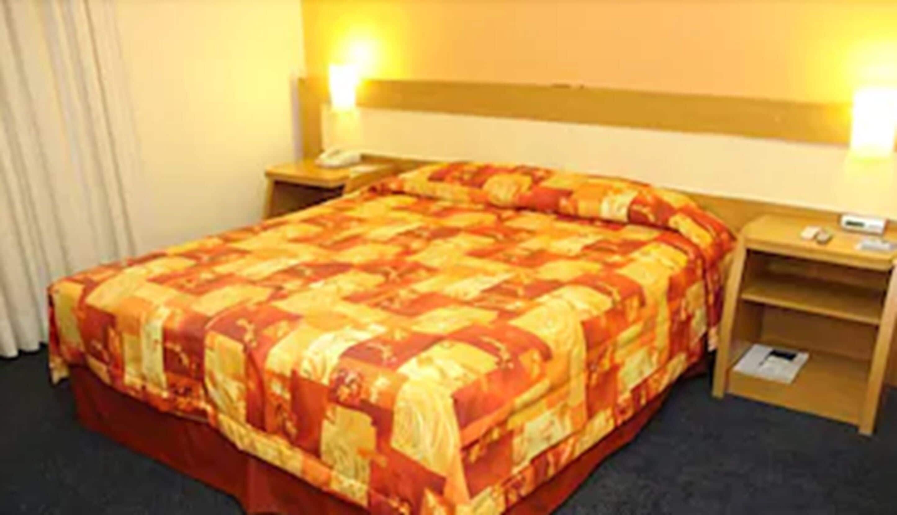 Quarto de hotel com uma cama grande, uma mesinha de cabeceira, uma luminária e um telefone.