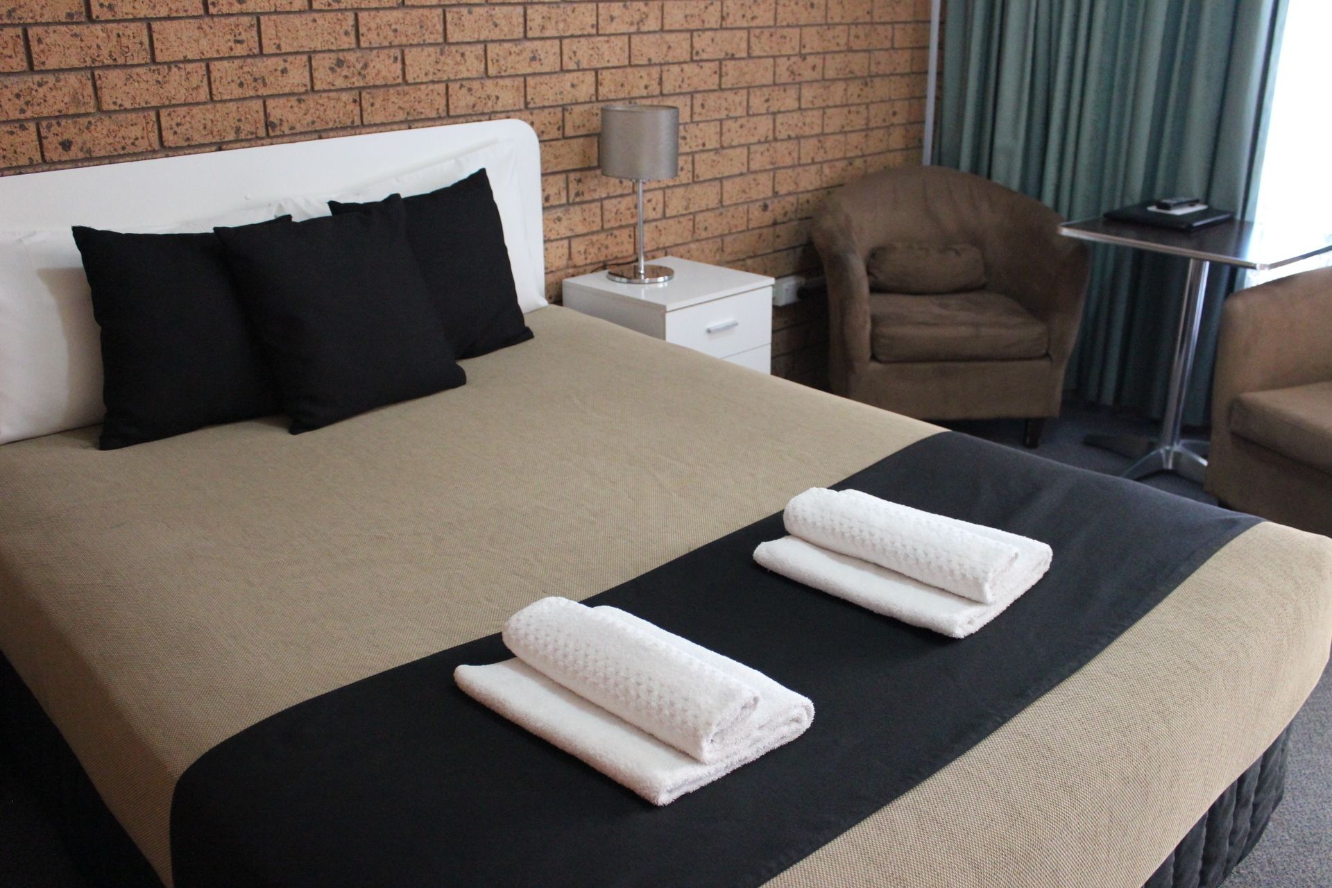 Uma cama bem arrumada com roupa de cama preta e bege, duas toalhas brancas, uma mesa de cabeceira branca, uma luminária e uma poltrona marrom em um quarto com paredes de tijolo.