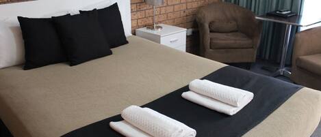 Uma cama bem arrumada com roupa de cama preta e bege, duas toalhas brancas, uma mesa de cabeceira branca, uma luminária e uma poltrona marrom em um quarto com paredes de tijolo.